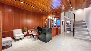 Uma área de recepção de hotel com balcão de mármore, painéis de parede em madeira, uma escadaria e um espaço de estar com cadeiras e um sofá.