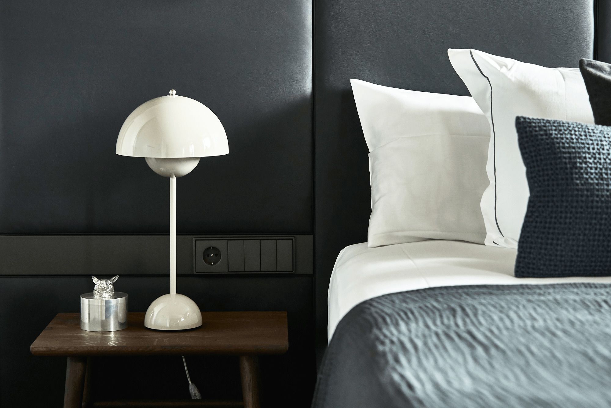 Cama bem arrumada com travesseiros brancos e azul-escuros, abajur branco em mesa de cabeceira de madeira e cabeceira escura.
