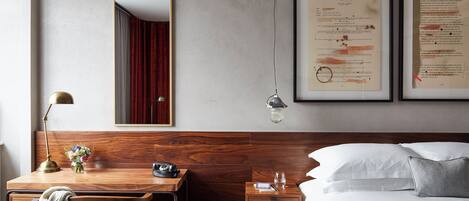 Quarto de hotel com cabeceira de madeira, escrivaninha com telefone e um vaso de flores, espelho e dois documentos emoldurados na parede.
