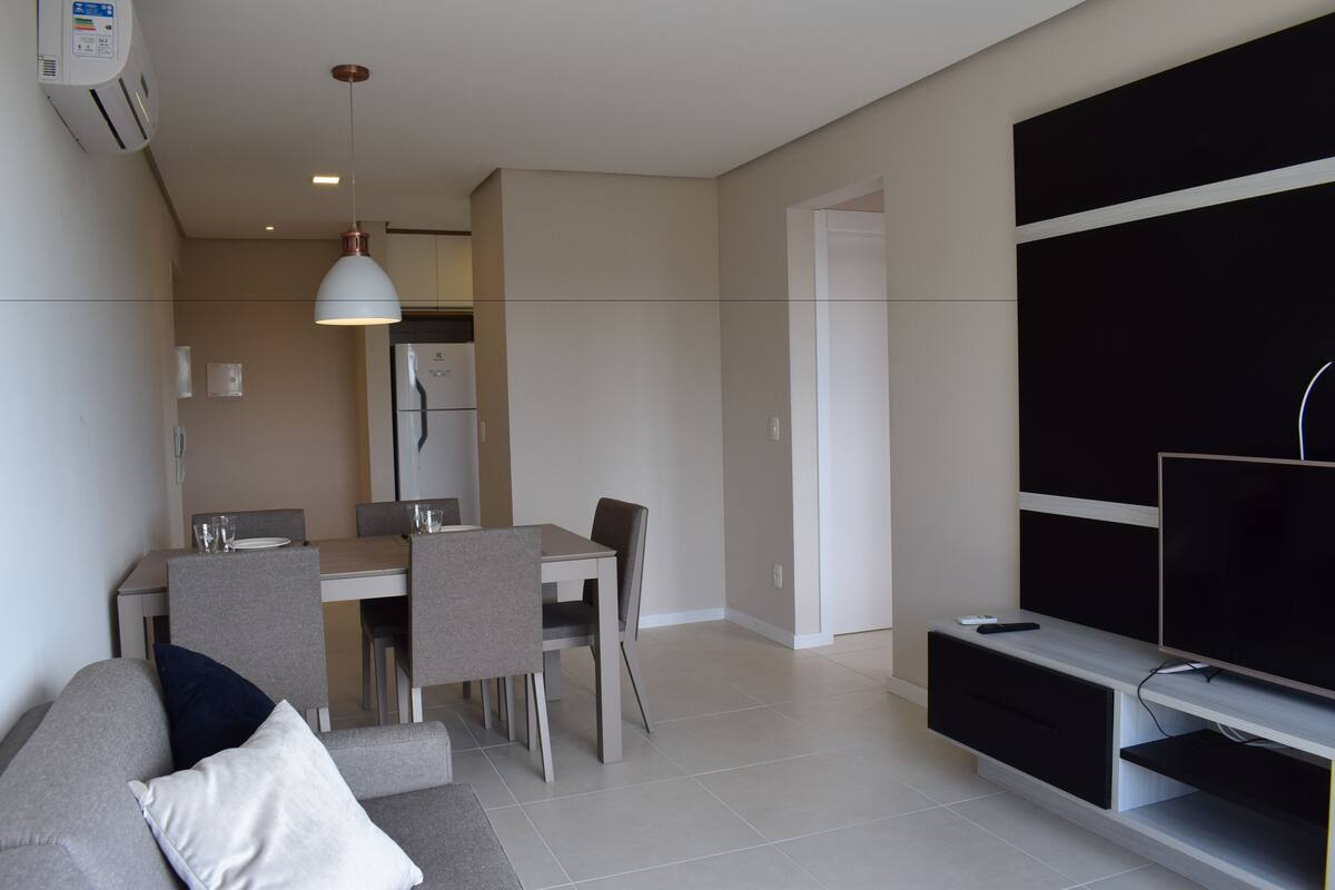 Uma sala de estar moderna com um sofá, uma mesa de jantar com cadeiras, uma TV em um suporte e uma cozinha ao fundo.