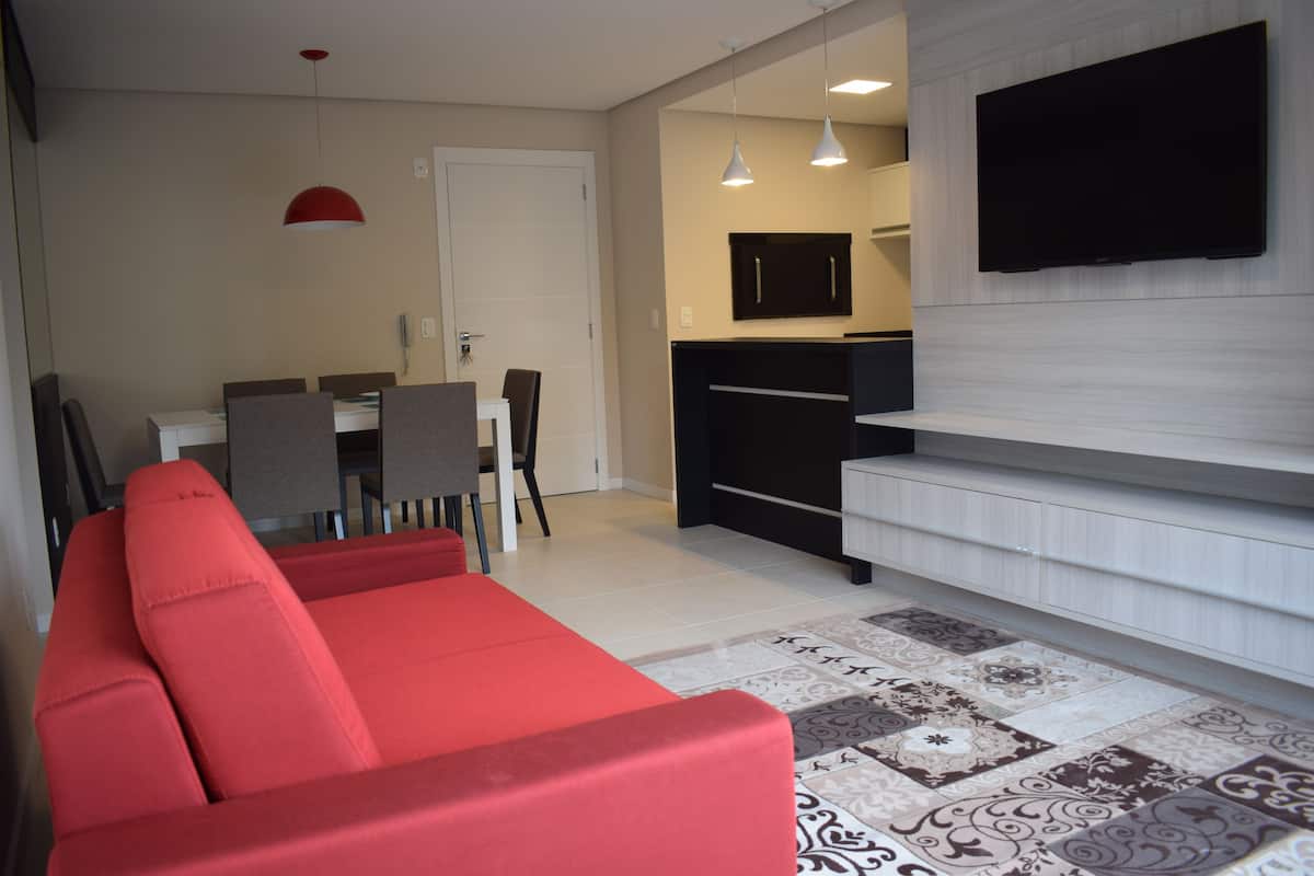 Uma sala de estar moderna com um sofá vermelho, um espaço de refeições com mesa e cadeiras, uma cozinha com uma TV fixada acima do balcão, e um tapete estampado no chão.