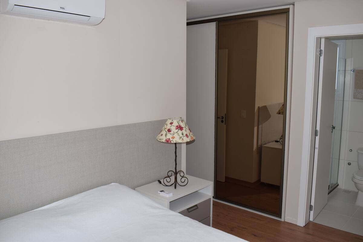 Quarto de hotel com cama, mesa de cabeceira com abajur, banheiro com chuveiro e porta que dá para outro quarto.