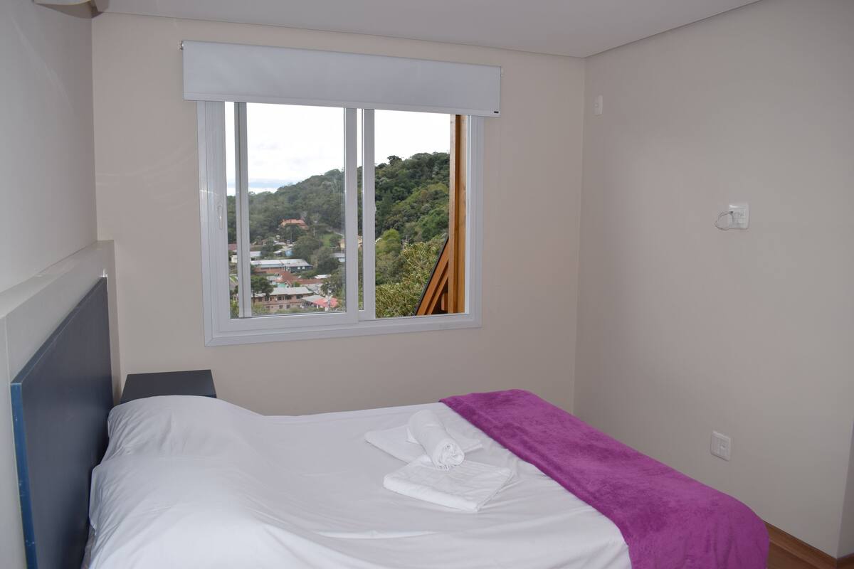 Uma cama de solteiro com colchão branco e manta roxa, uma janela com vista para uma paisagem verde, e uma parede branca com um interruptor de luz.