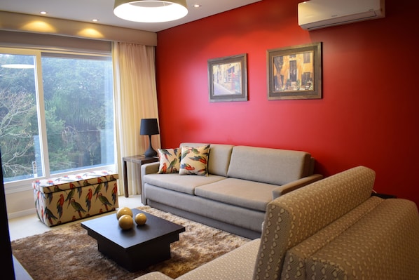 Uma sala de estar com parede vermelha, sofá cinza, um banquinho estampado e uma mesa de centro com bolas decorativas.