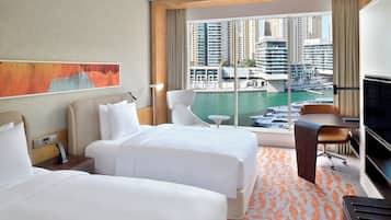 Quarto de hotel com duas camas, uma mesa, televisão e uma grande janela com vista para uma marina e edifícios.
