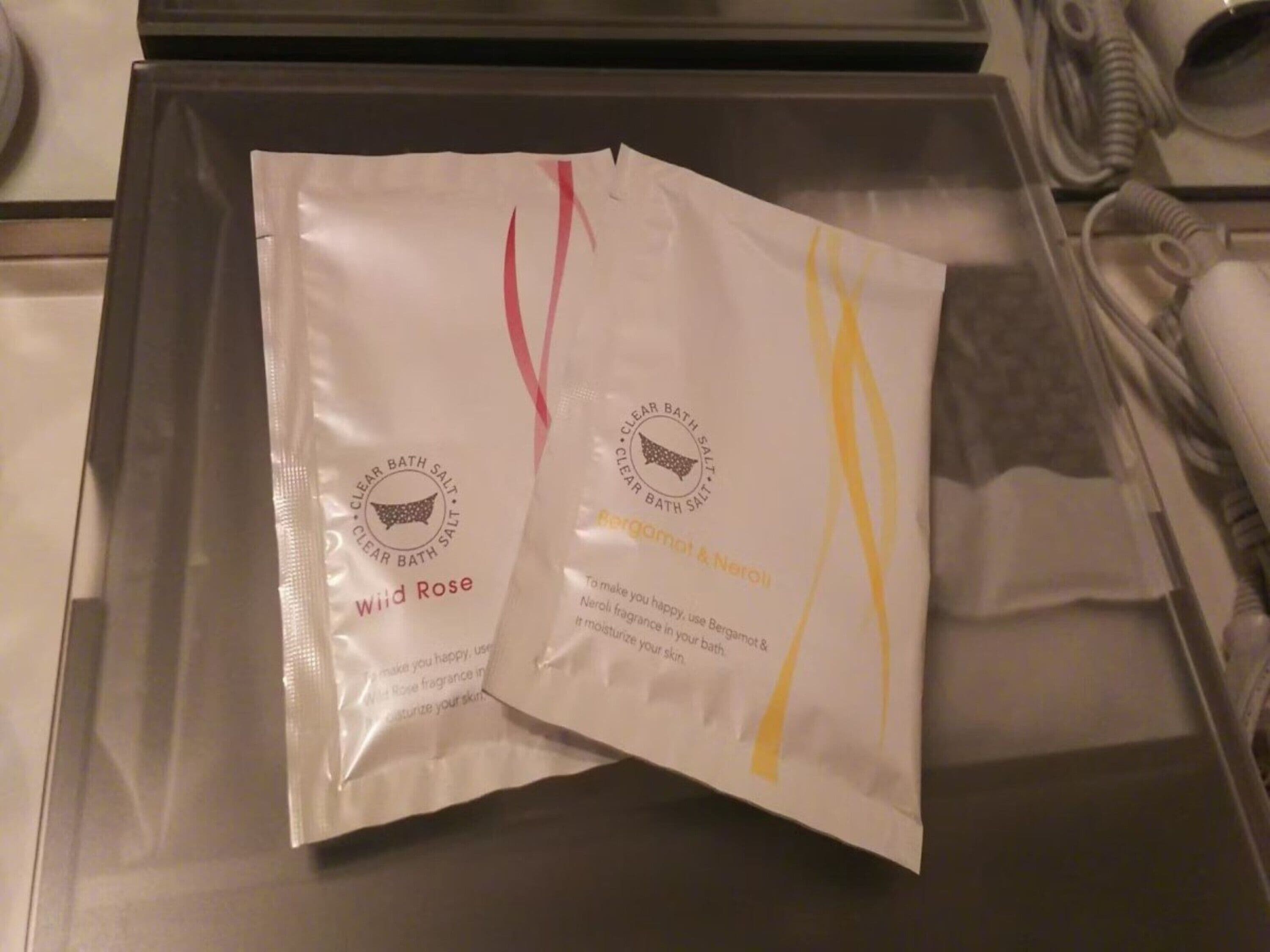 Dois saquinhos de sais de banho transparentes, um identificado como 'Wild Rose' e outro como 'Bergamota & Neroli', sobre uma bandeja.