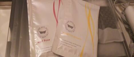 Dois saquinhos de sais de banho transparentes, um identificado como 'Wild Rose' e outro como 'Bergamota & Neroli', sobre uma bandeja.