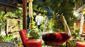 Uma mão segurando uma taça de vinho com líquido vermelho, uma cadeira vermelha e uma mesa de madeira em ambiente externo com vegetação e uma estrutura de madeira ao fundo.