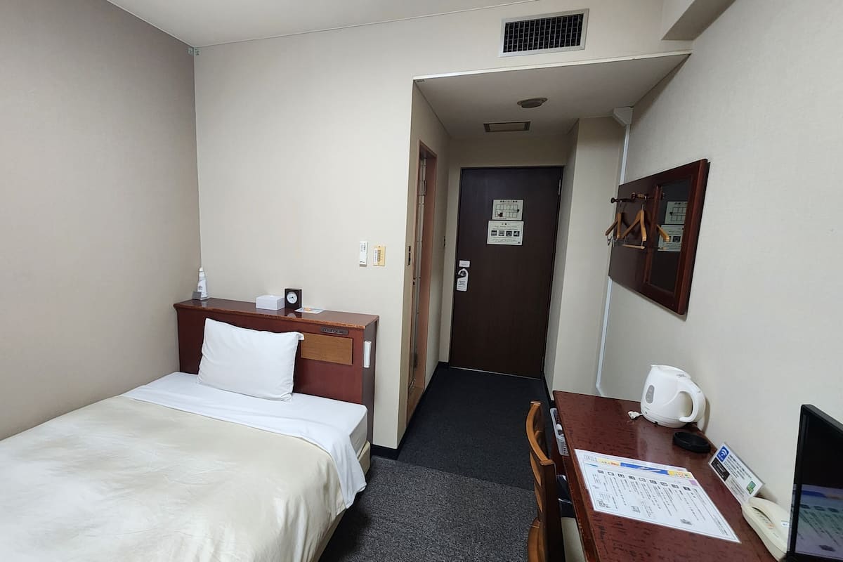 Quarto de hotel com cama, escrivaninha, cadeira, cafeteira e porta que dá para outro cômodo.