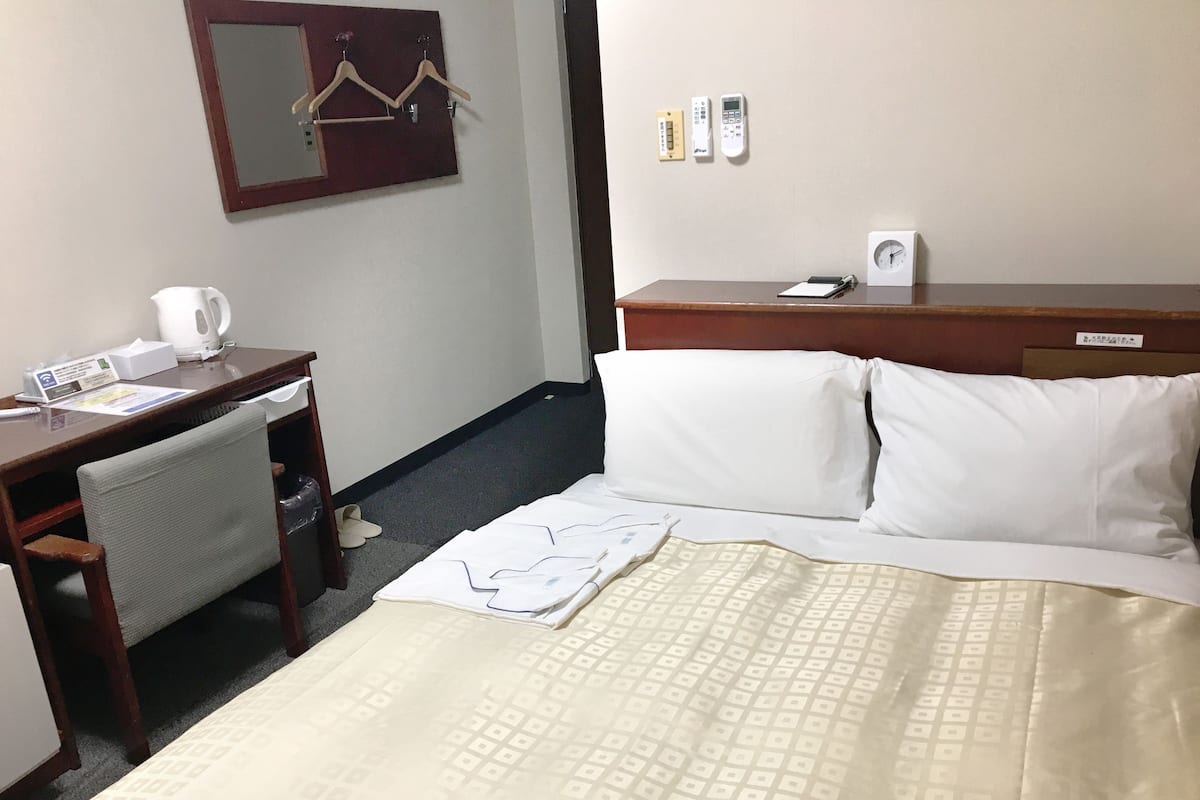 Quarto de hotel com cama, escrivaninha com cadeira, espelho, relógio e termostato na parede.