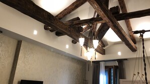 Quarto com vigas de madeira aparentes, um lustre, uma TV de tela plana na parede e uma janela com cortinas.