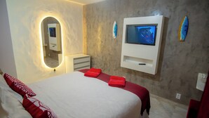 Quarto de hotel com cama, televisão fixada na parede, espelho e uma decoração com prancha de surf.