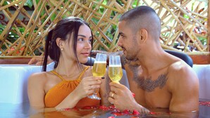 Duas pessoas brindando com bebidas em uma banheira de hidromassagem cercada por vegetação.