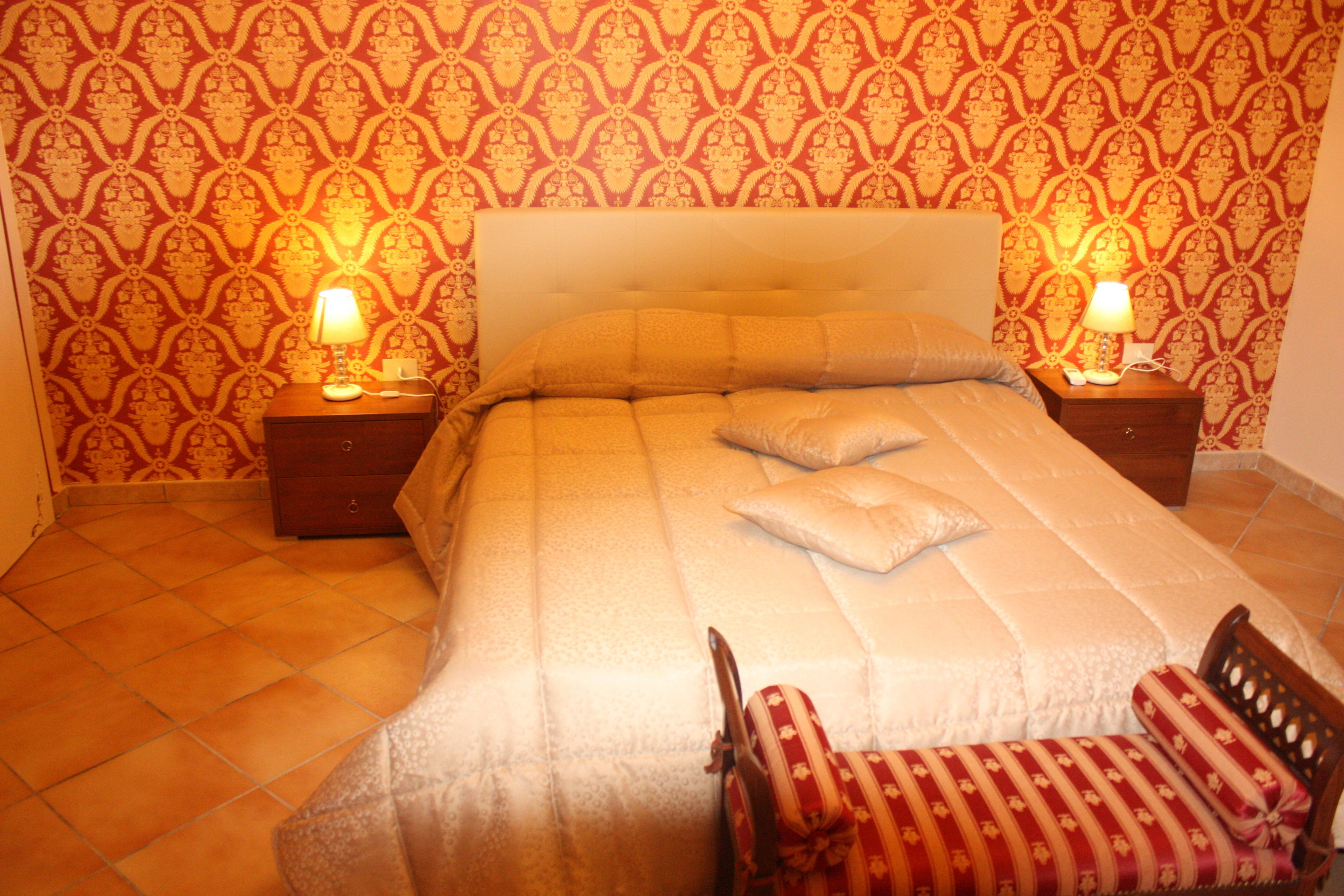 Uma cama bem arrumada com edredom branco e duas almofadas, com duas mesas de cabeceira com abajures, num quarto com papel de parede estampado e chão de azulejos.