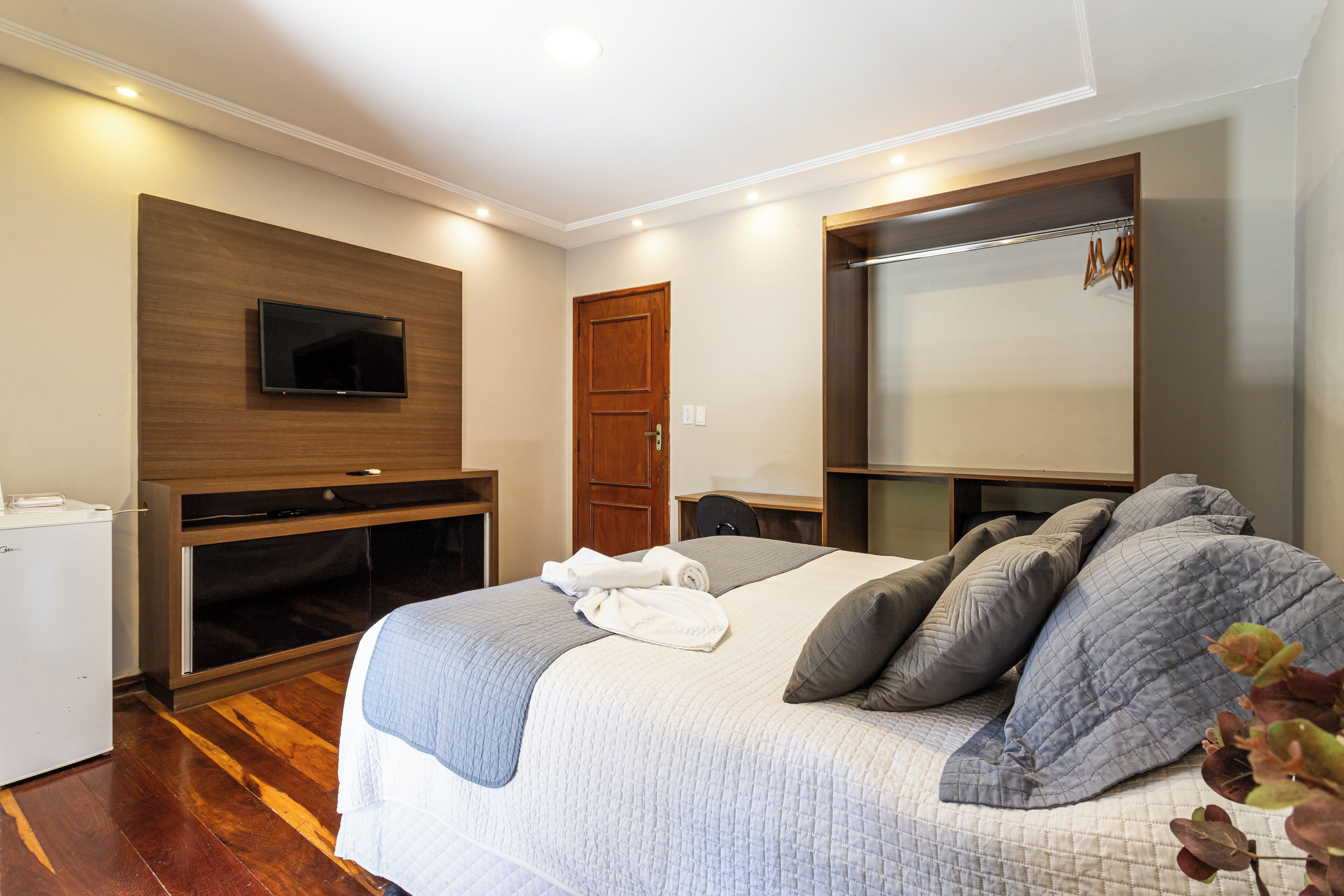 Quarto moderno com cama, televisão fixada em painel de madeira, guarda-roupa e espelho.