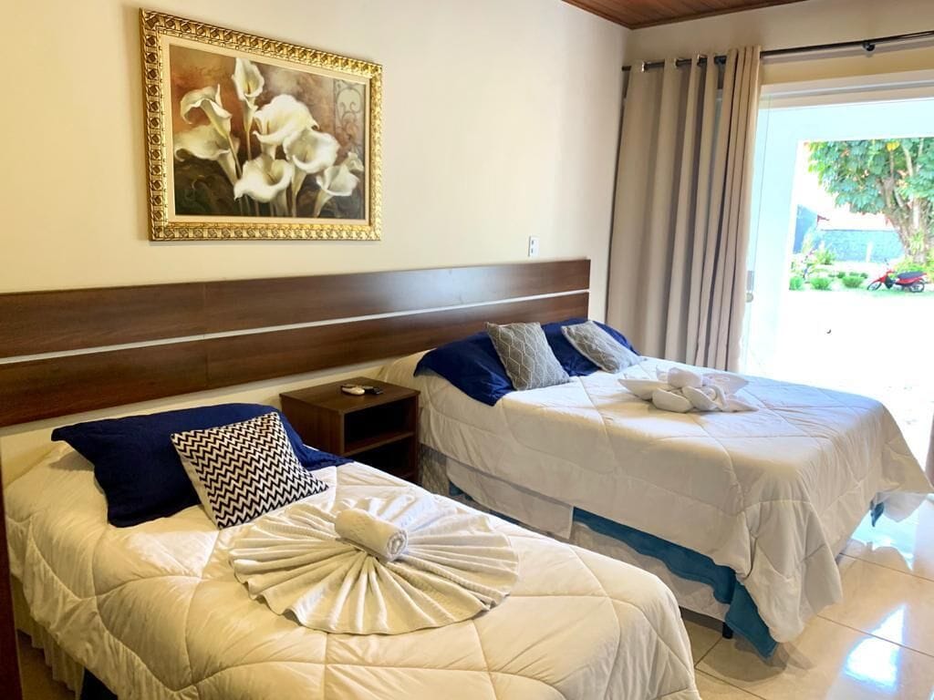 Quarto de hotel com duas camas, cabeceira de madeira, um quadro na parede e vista para o exterior pela janela.