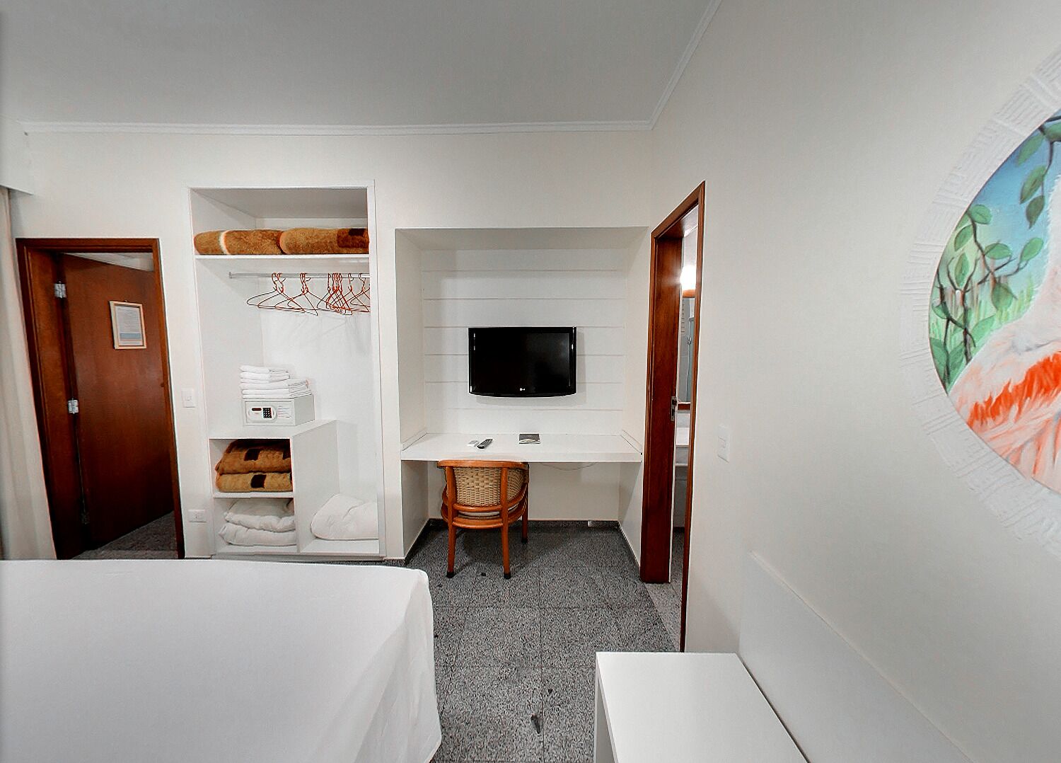 Quarto de hotel com uma cama, uma escrivaninha com cadeira, televisão e guarda-roupa.