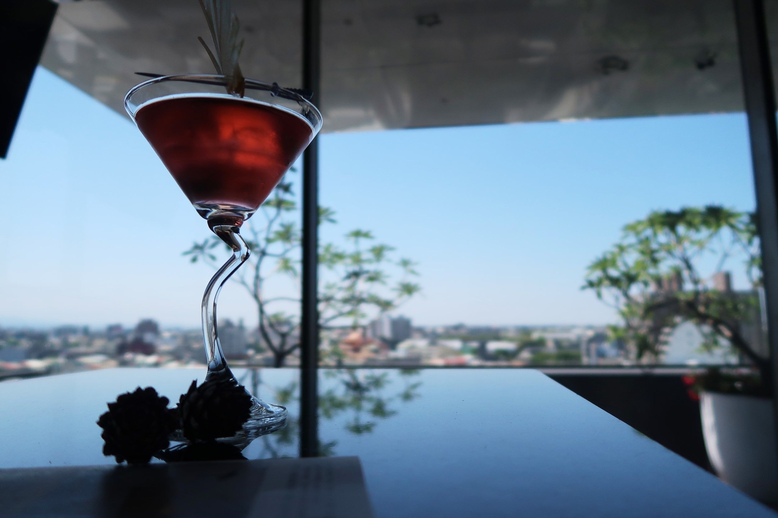 Um copo de martini com uma bebida vermelha, colocado sobre uma mesa com vista para a cidade ao fundo.