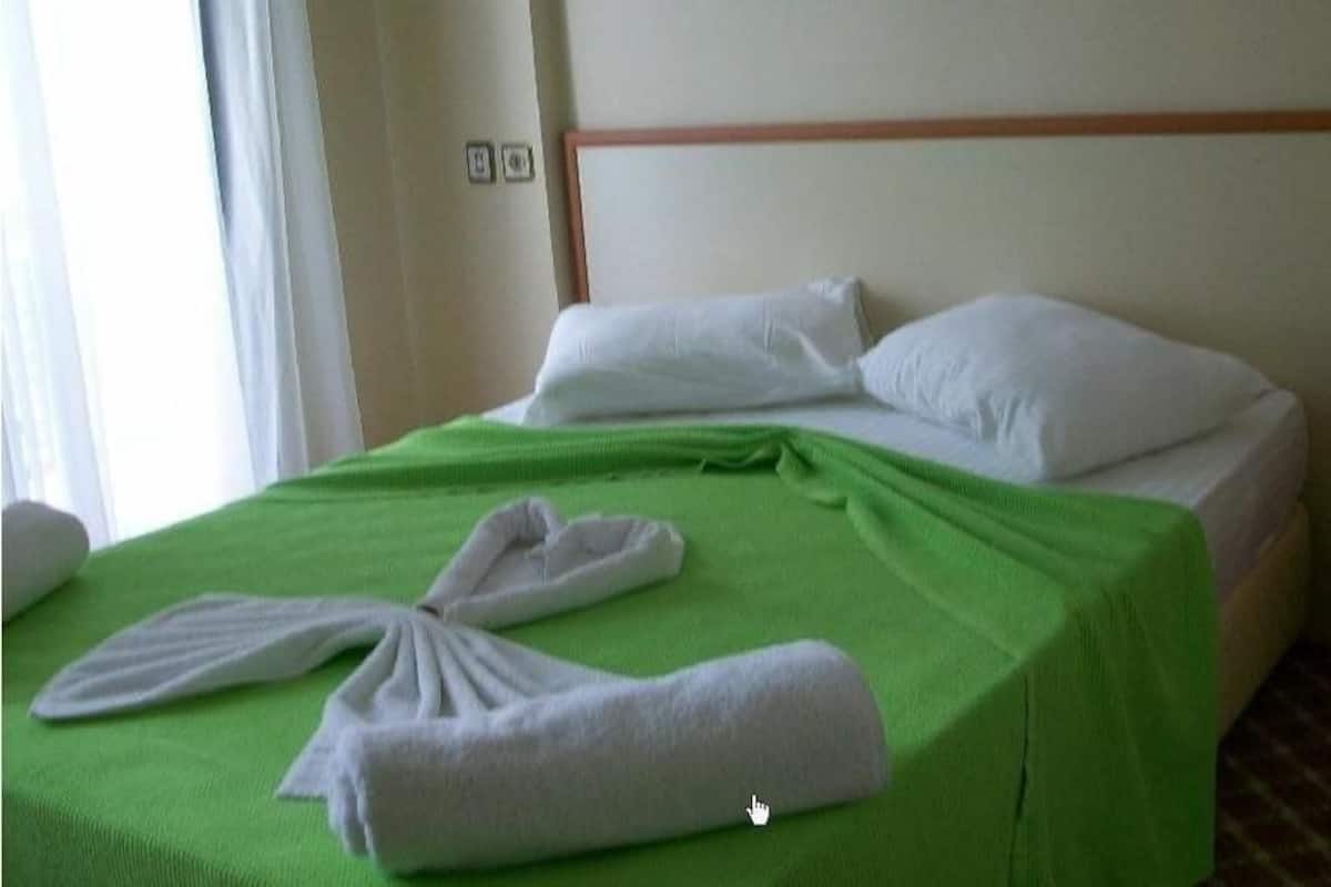 Quarto de hotel com cama, colcha verde, almofadas brancas e duas toalhas brancas dobradas na cama.
