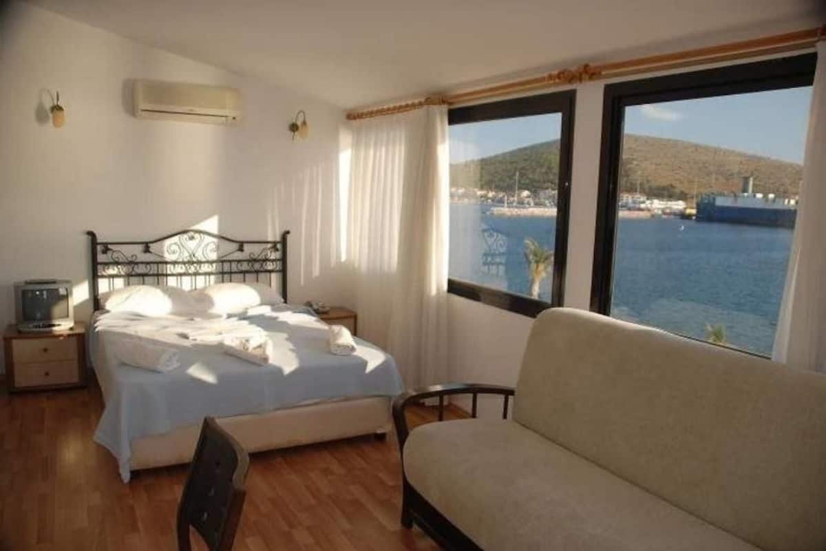 Quarto com cama, cadeira, mesa de cabeceira e janela com vista para um corpo d'água e uma formação terrestre ao longe.