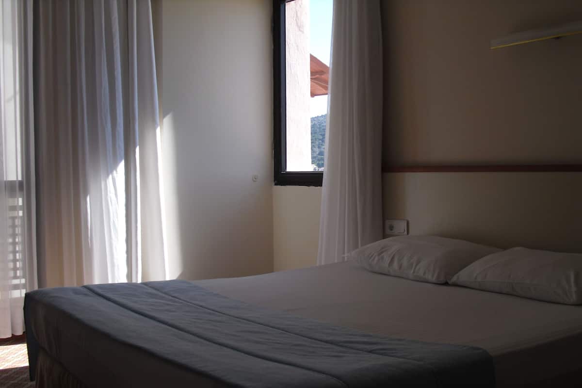 Quarto de hotel com uma cama grande, cortinas brancas e janela com vista para um edifício.