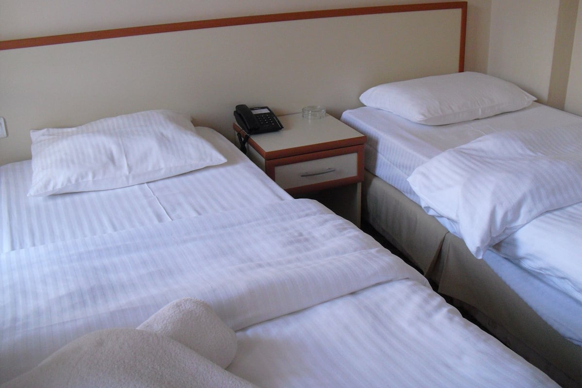 Quarto de hotel com duas camas de solteiro, uma mesinha de cabeceira com telefone e uma gaveta pequena.