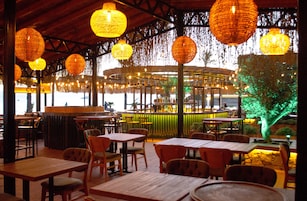 Um restaurante interno com mesas e cadeiras de madeira, lanternas penduradas e uma árvore com iluminação verde.