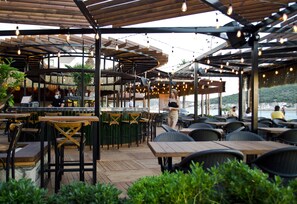 Restaurante ao ar livre com mesas e cadeiras de madeira, área de bar e luzes de corda.