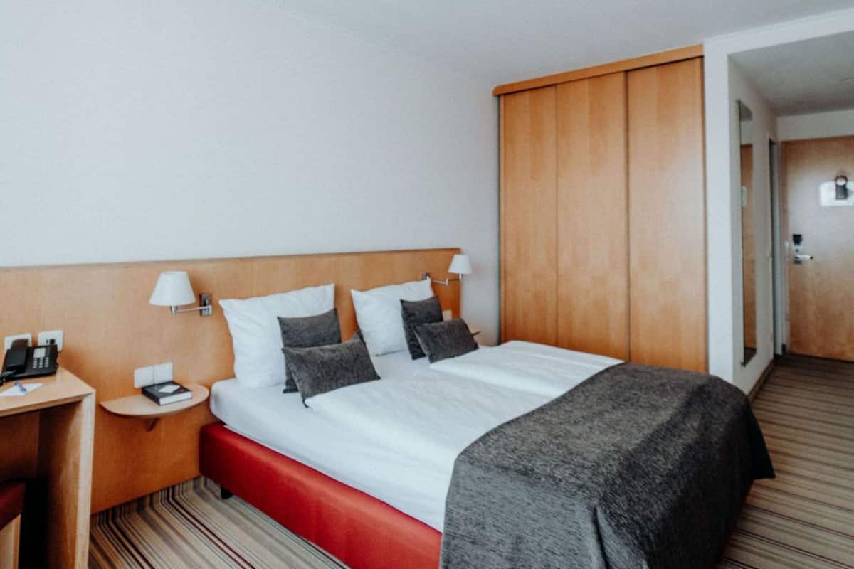 Quarto de hotel com cama, escrivaninha, telefone, abajur e porta.