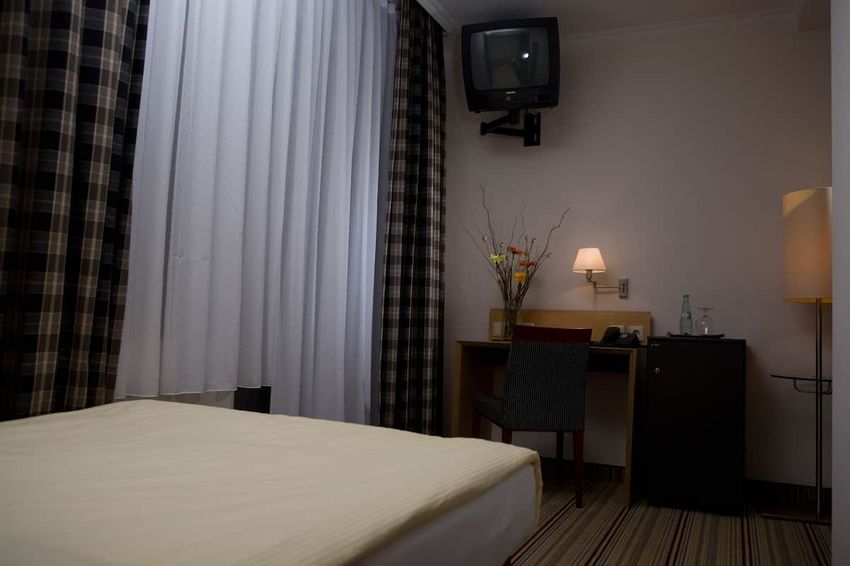 Quarto de hotel com cama, escrivaninha com cadeira, televisão fixada na parede e luminária.