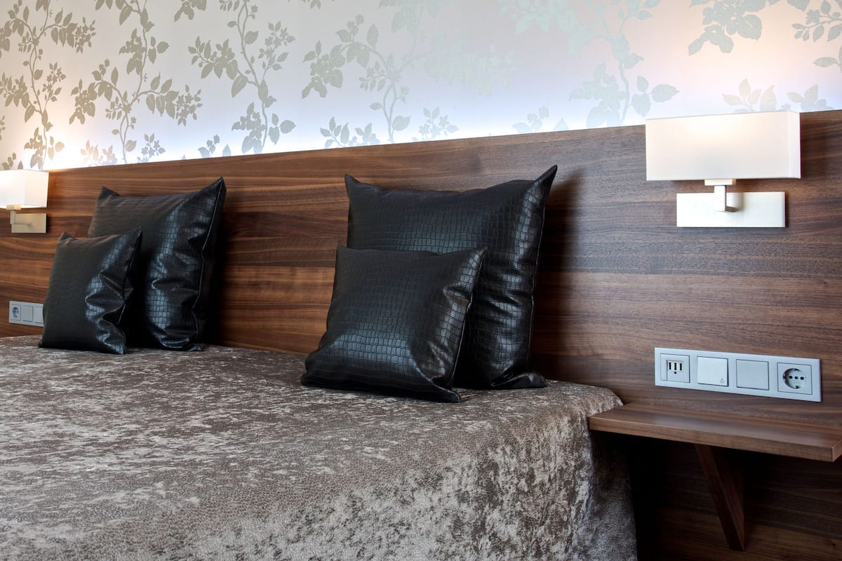 Quarto de hotel com cabeceira de madeira, duas almofadas pretas, cama com manta texturizada, mesa de cabeceira com abajur e parede com papel de parede floral.