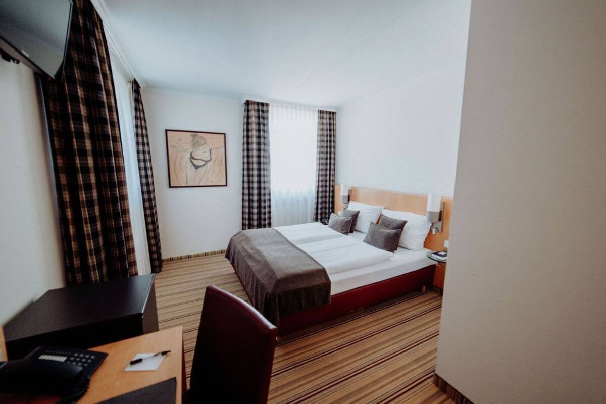Quarto de hotel com cama, escrivaninha, cadeira, televisão e um quadro na parede.