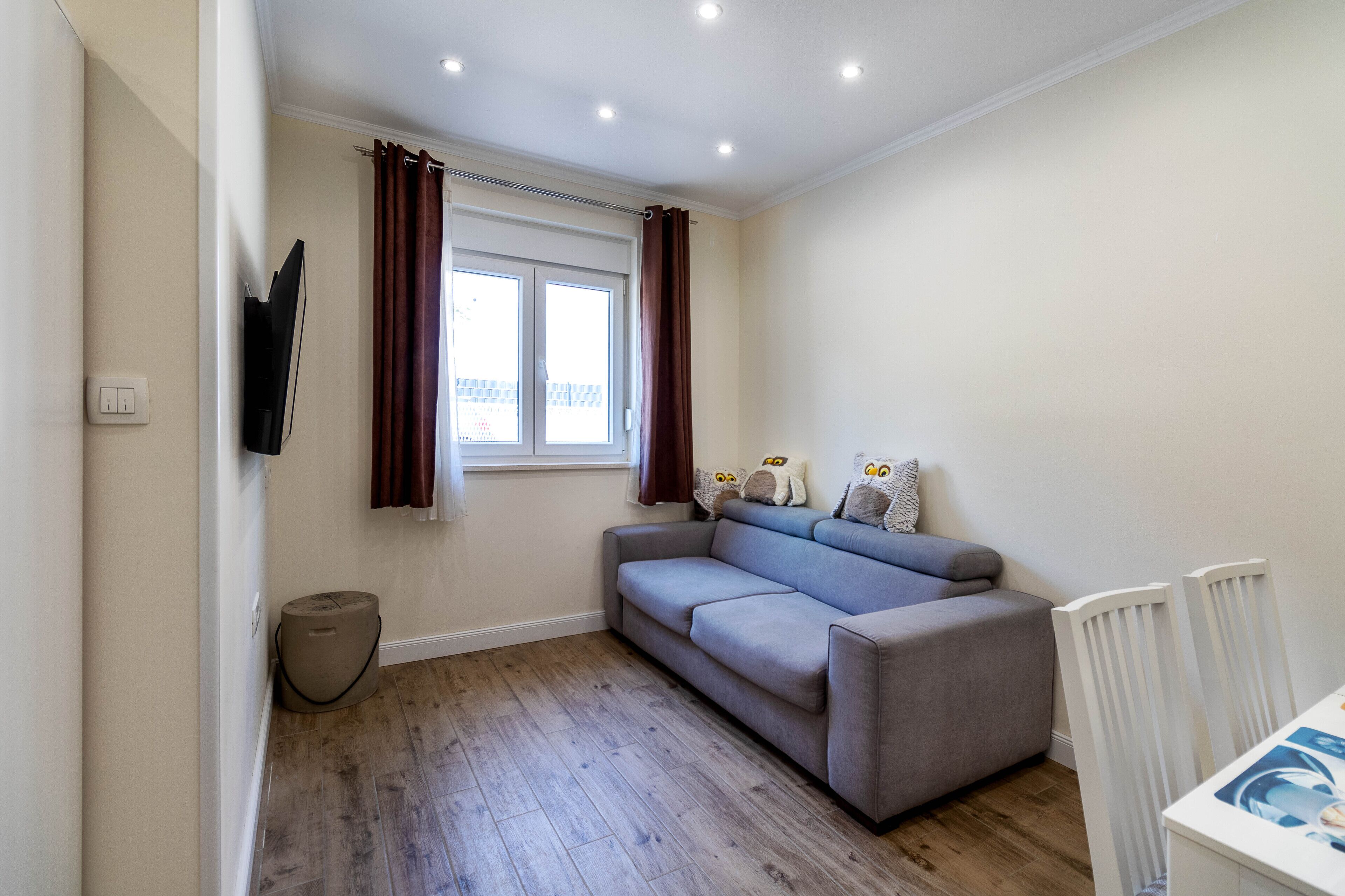 Quarto com sofá cinzento, janela com cortinas, radiador e um banquinho redondo pequeno.