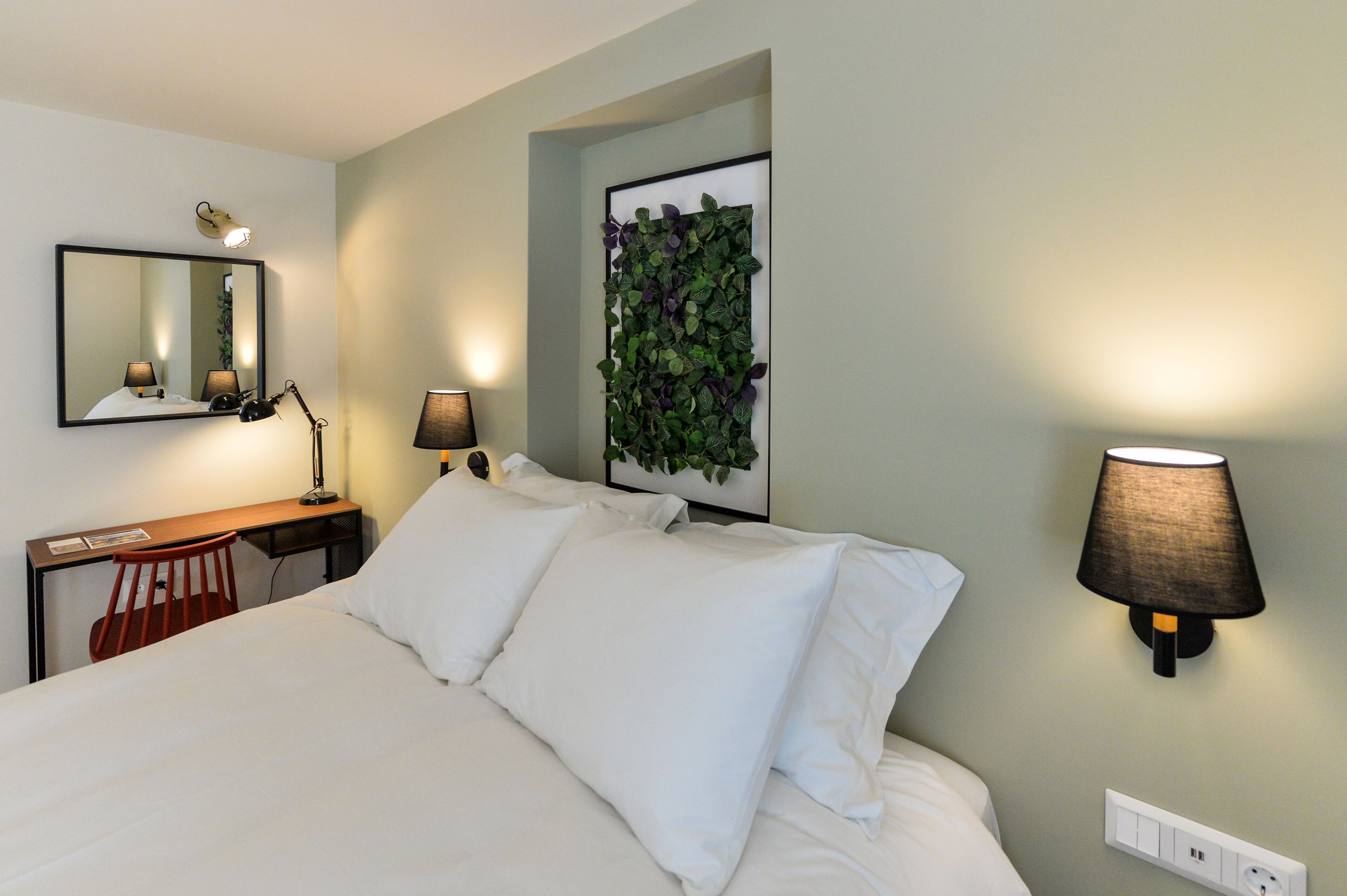 Quarto de hotel com cama, escrivaninha, cadeira, abajur e planta fixada na parede.