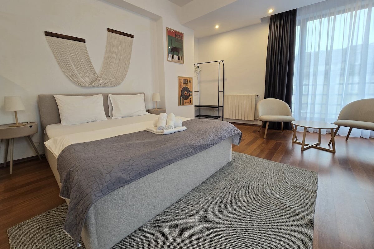 Quarto de hotel moderno com uma cama grande, área de estar com uma poltrona e uma mesa pequena, radiador e janela com cortinas.