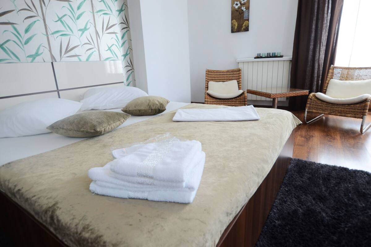Uma cama bem arrumada com lençóis brancos e uma toalha dobrada, uma cadeira de vime e uma pequena mesa com uma garrafa e uma xícara.