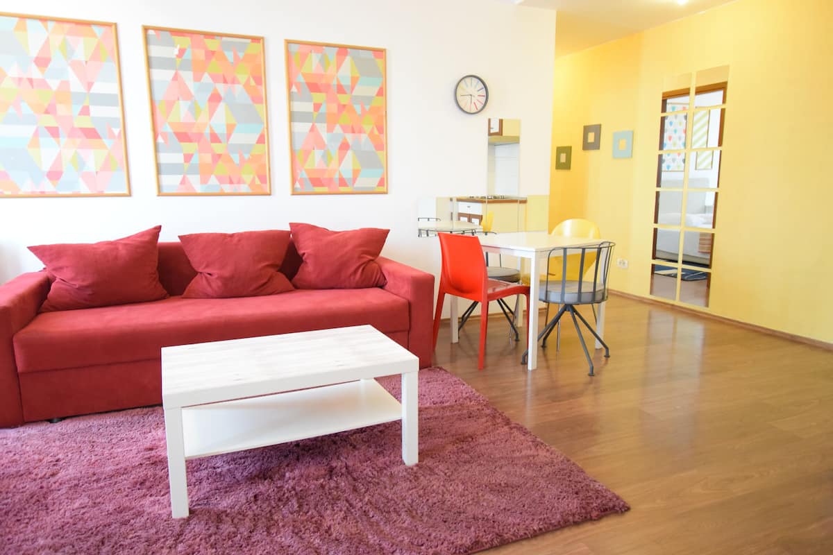 Uma sala de estar com um sofá vermelho, uma mesa de centro branca e um tapete roxo. Há quadros abstratos na parede, um relógio e um espaço de jantar com uma mesa branca e cadeiras laranjas.