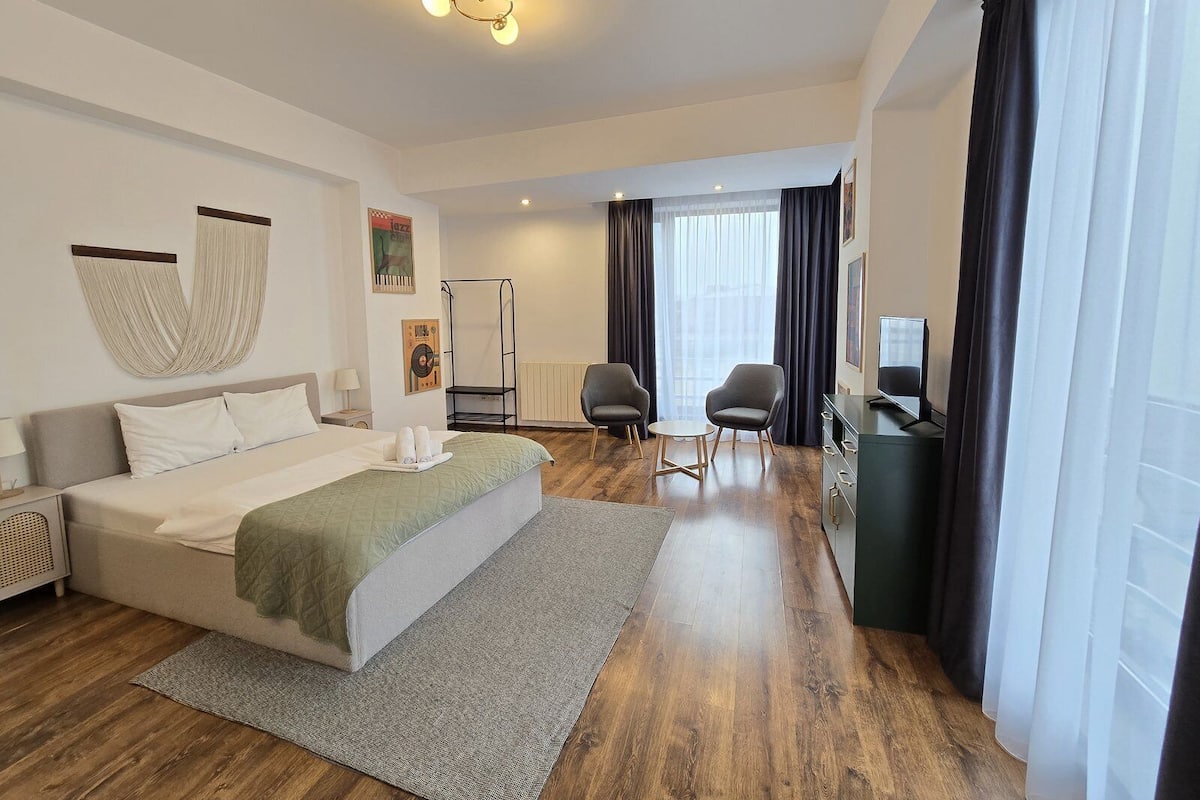 Quarto de hotel moderno com uma cama grande, uma escrivaninha, duas cadeiras, uma TV e uma janela com cortinas.