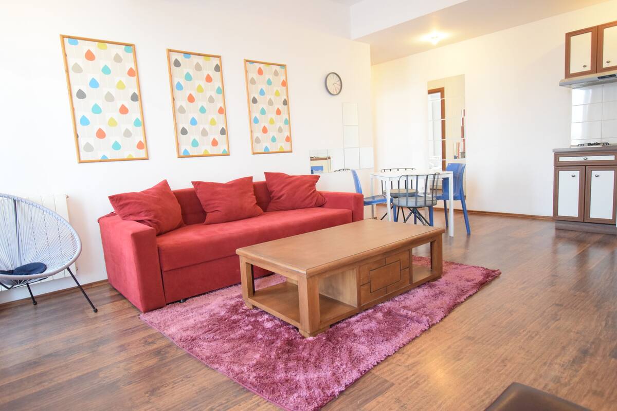 Sala de estar com um sofá vermelho, uma mesa de centro de madeira e um tapete rosa. Há três obras de arte emolduradas na parede, um relógio e uma área de cozinha ao fundo.