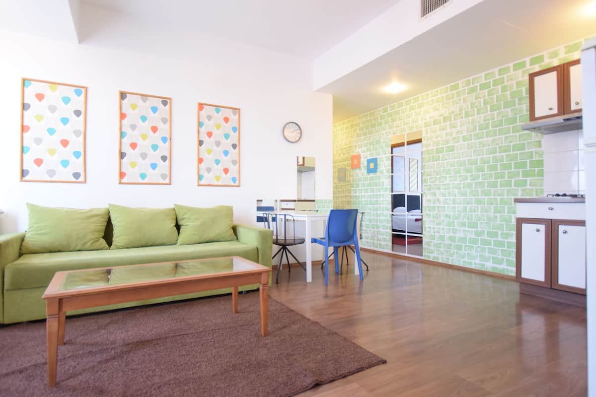 Sala de estar com sofá verde, mesa de centro de madeira e tapete marrom. Há uma área de jantar com mesa e cadeiras brancas, e uma cozinha com paredes de tijolos verdes e armários brancos.