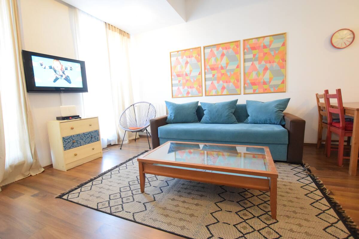 Uma sala de estar com um sofá azul, uma mesa de centro de vidro, uma TV na parede e um tapete estampado.