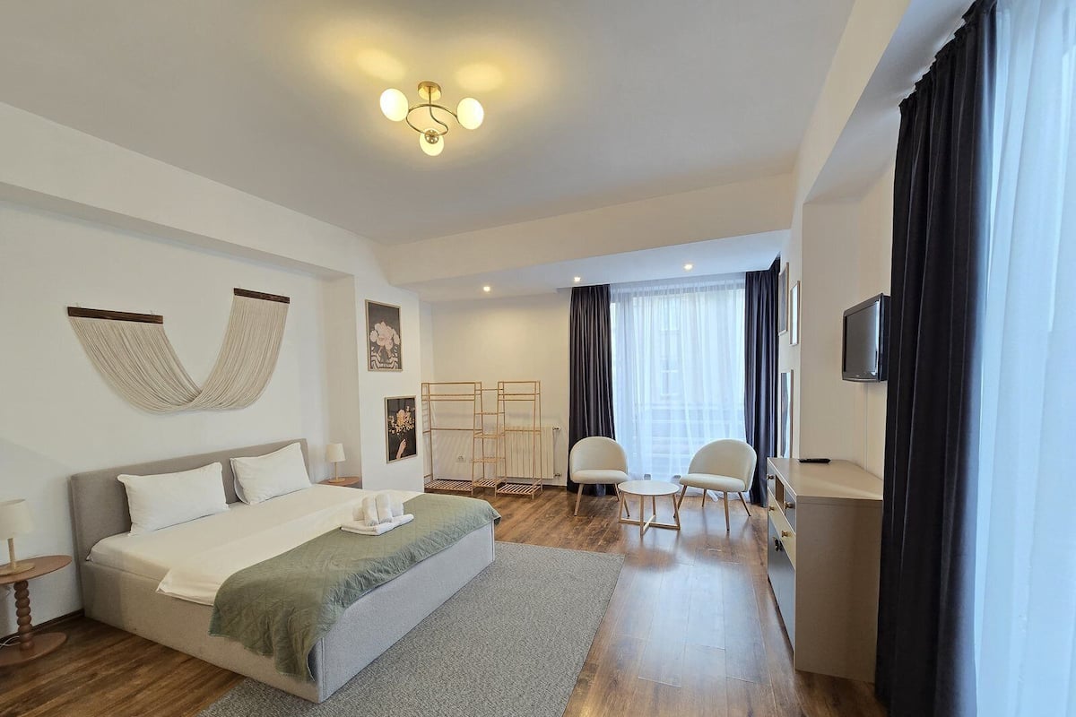 Quarto de hotel moderno com uma cama grande, uma escrivaninha com duas cadeiras, uma televisão e uma janela ampla com cortinas.