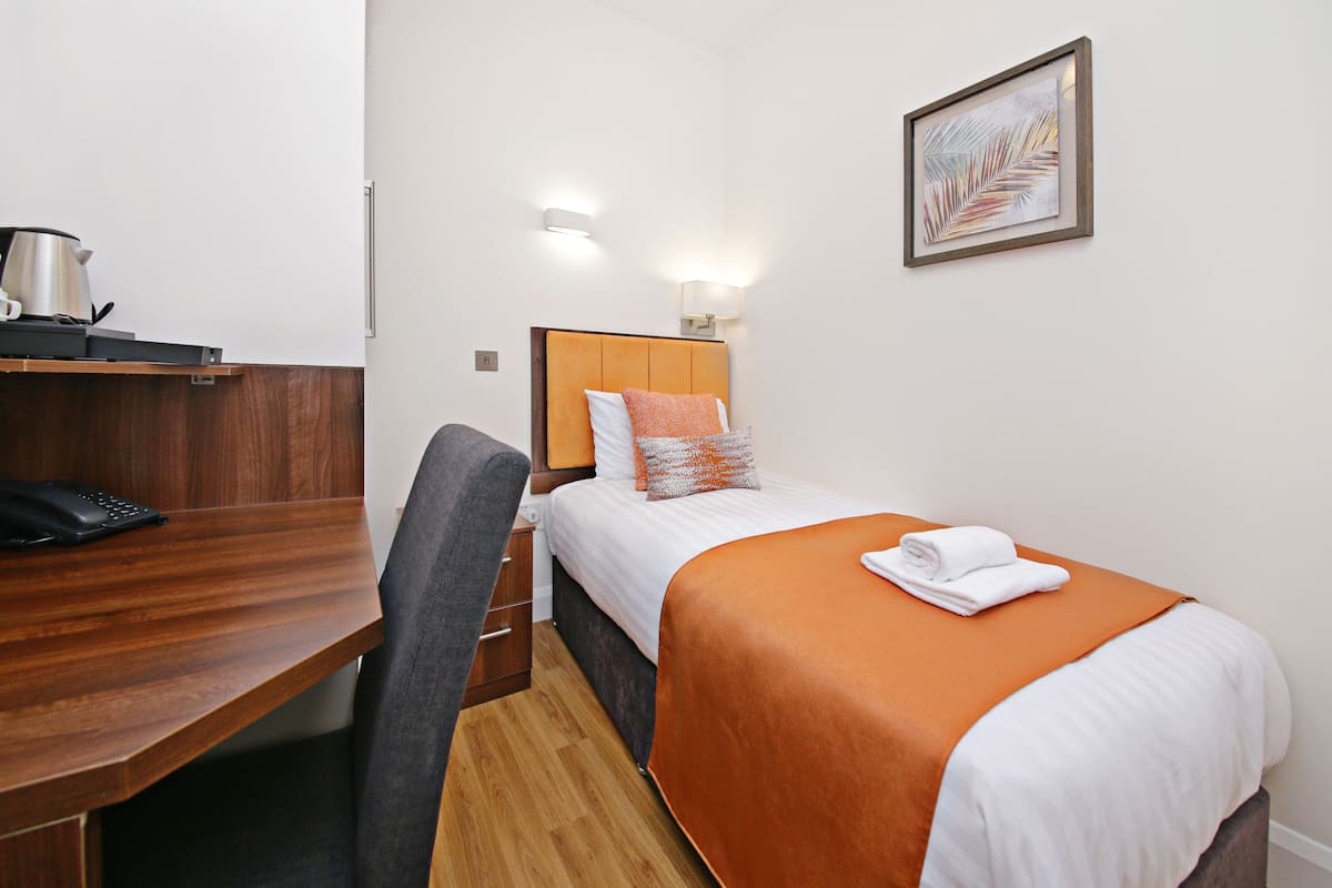 Quarto de hotel com escrivaninha de madeira, cama com manta laranja, cadeira cinza e quadro na parede.
