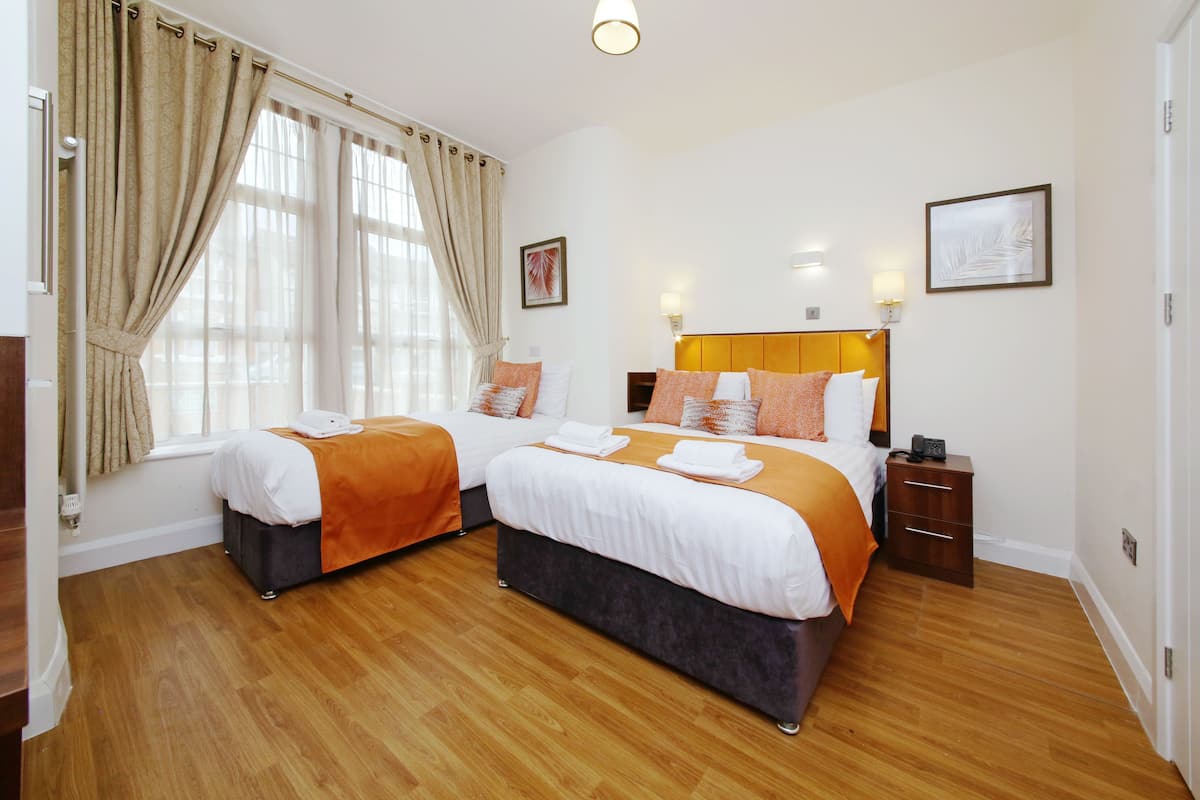 Quarto de hotel com duas camas, piso de madeira e uma janela grande com cortinas.