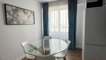 Uma cozinha moderna com geladeira branca, mesa de jantar de vidro e quatro cadeiras, e uma janela com cortinas azuis.