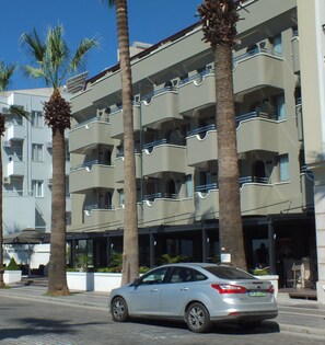 Um hotel com vários andares, varandas, palmeiras e um carro estacionado na frente.