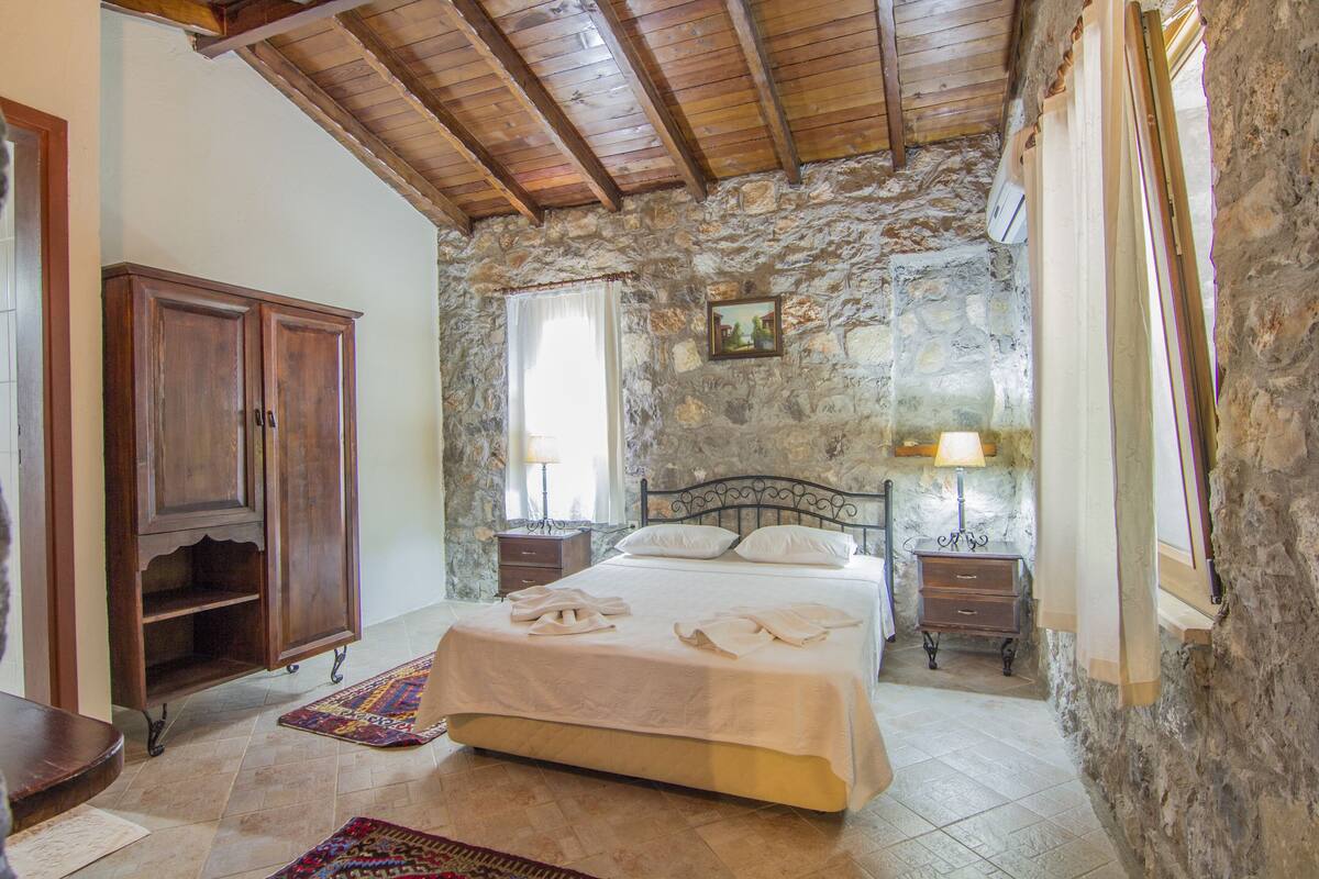 Quarto com parede de pedra, teto de madeira, cama com roupa de cama branca, guarda-roupa de madeira, mesa de cabeceira com abajur e janela com cortinas.