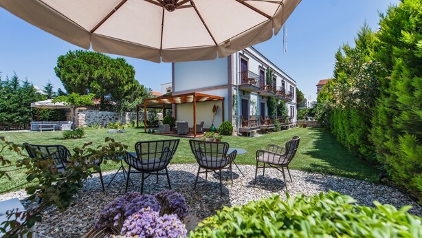 Um pátio externo com um guarda-sol grande, mesas e cadeiras ao ar livre, e um jardim bem cuidado.
