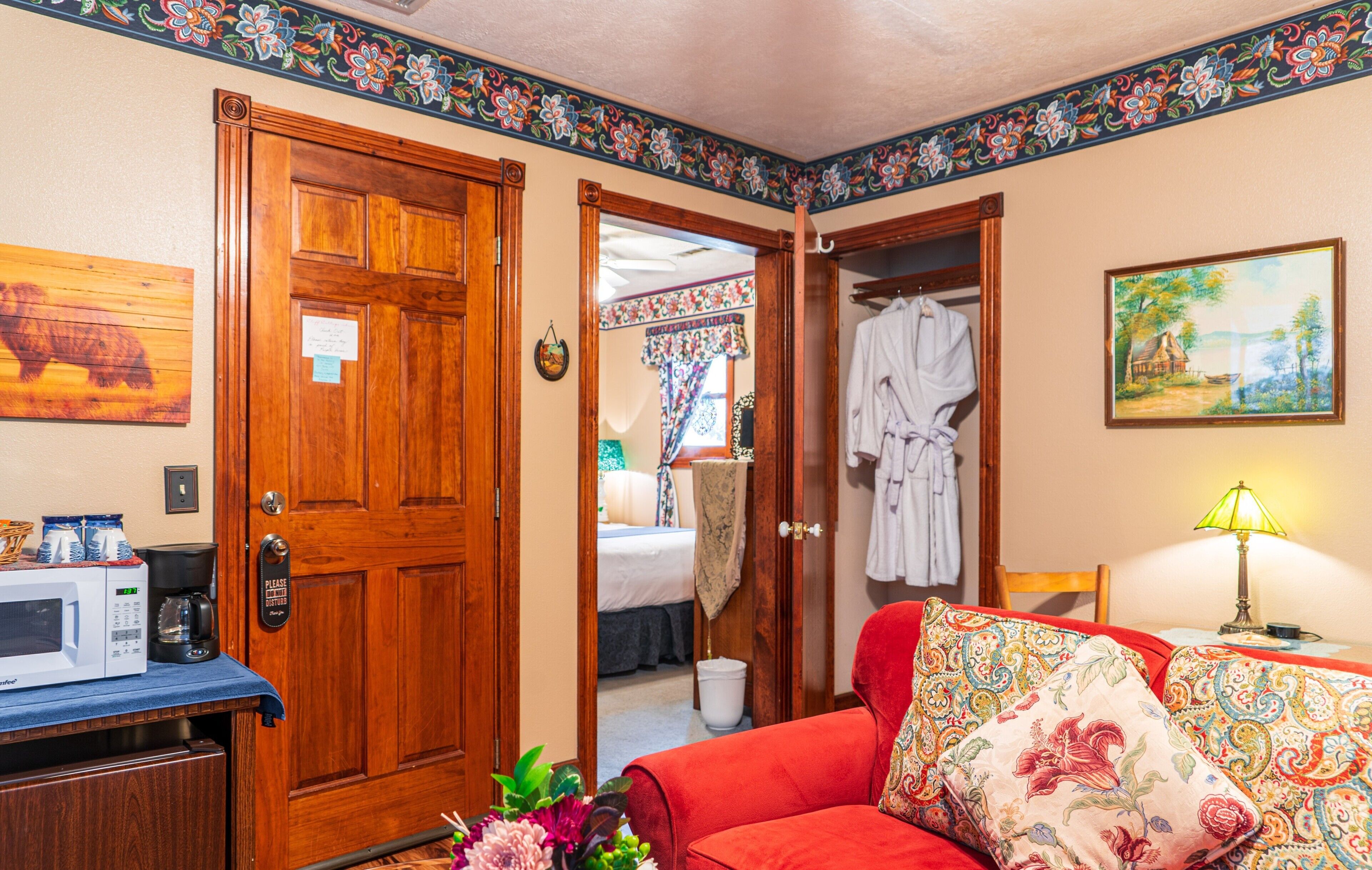 Quarto de hotel com porta de madeira, uma cama, sofá vermelho, um quadro, uma lâmpada e micro-ondas no balcão.