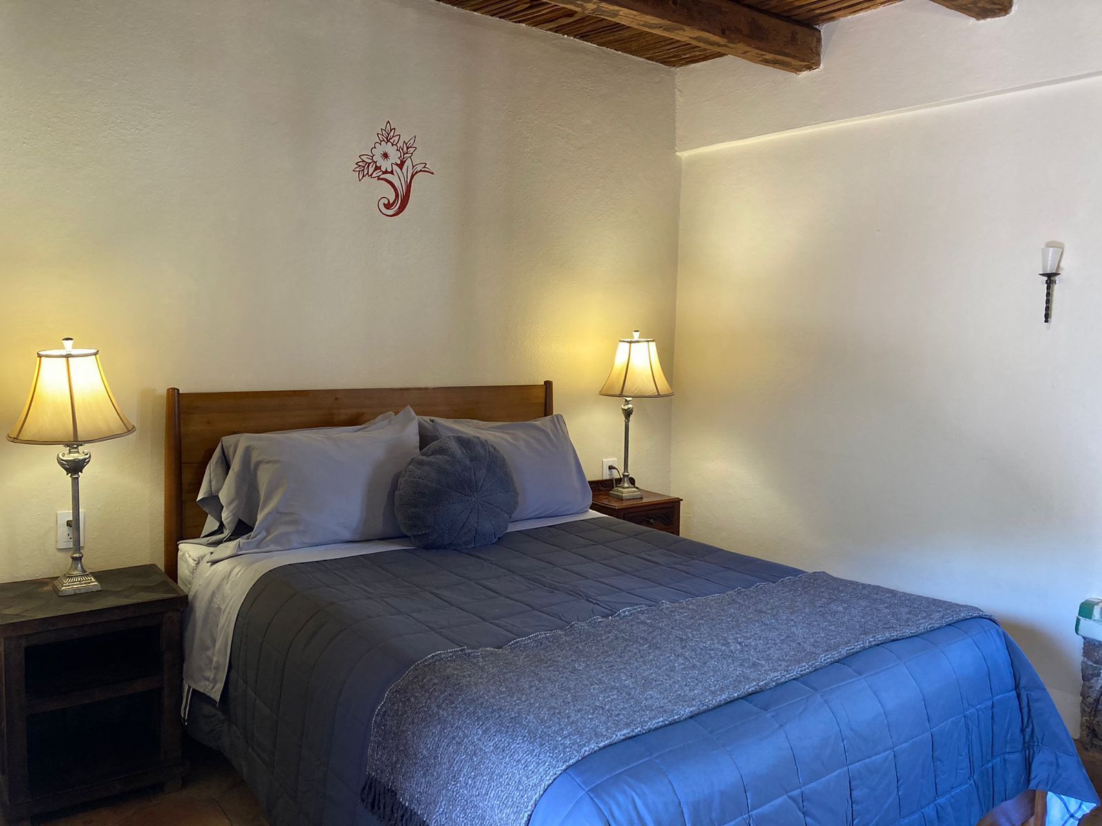 Quarto com cama, duas luminárias de cabeceira, um detalhe decorativo na parede e uma viga de madeira no teto.